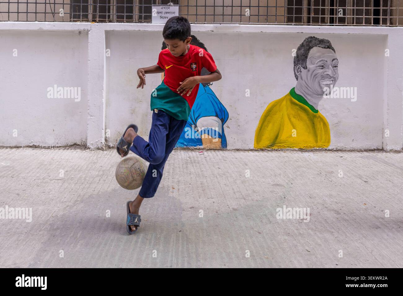 A boy seen playing foot ball in front of a graffiti of famous football ...