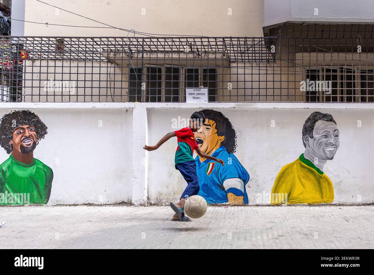 A boy seen playing foot ball in front of a graffiti of famous football ...
