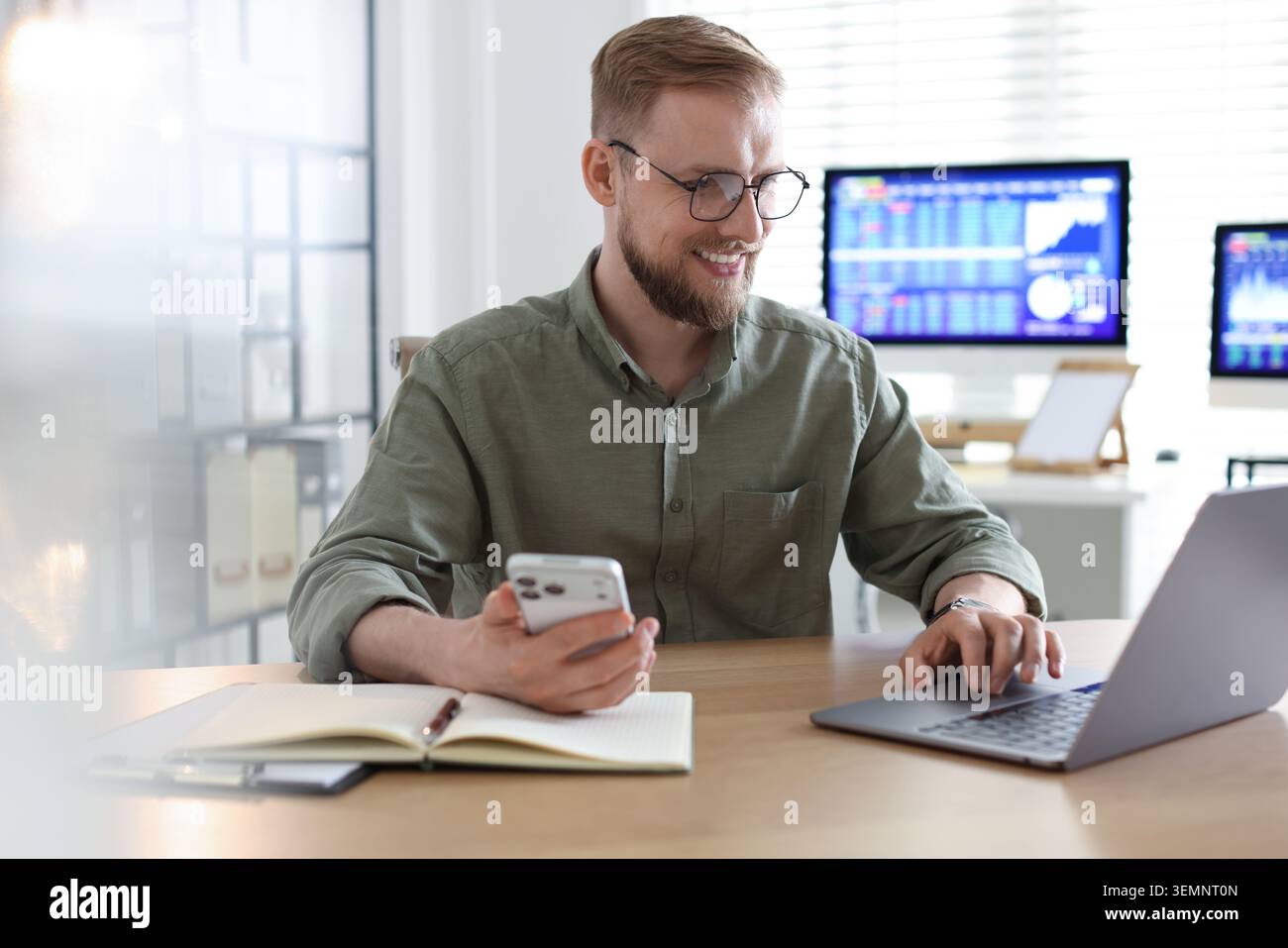 Financial trading specialist with smartphone working on laptop at desk ...