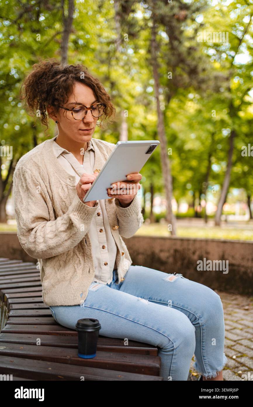 Adult woman working on a tablet outdoors while sitting on a bench in a ...