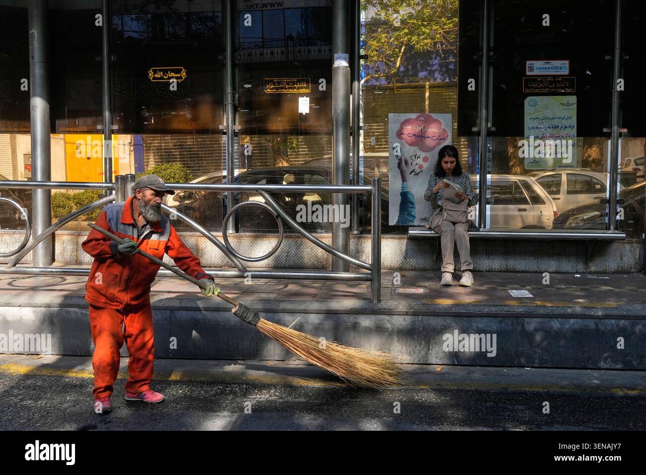 A woman sits at a bus stop as a city worker sweeps the street in ...