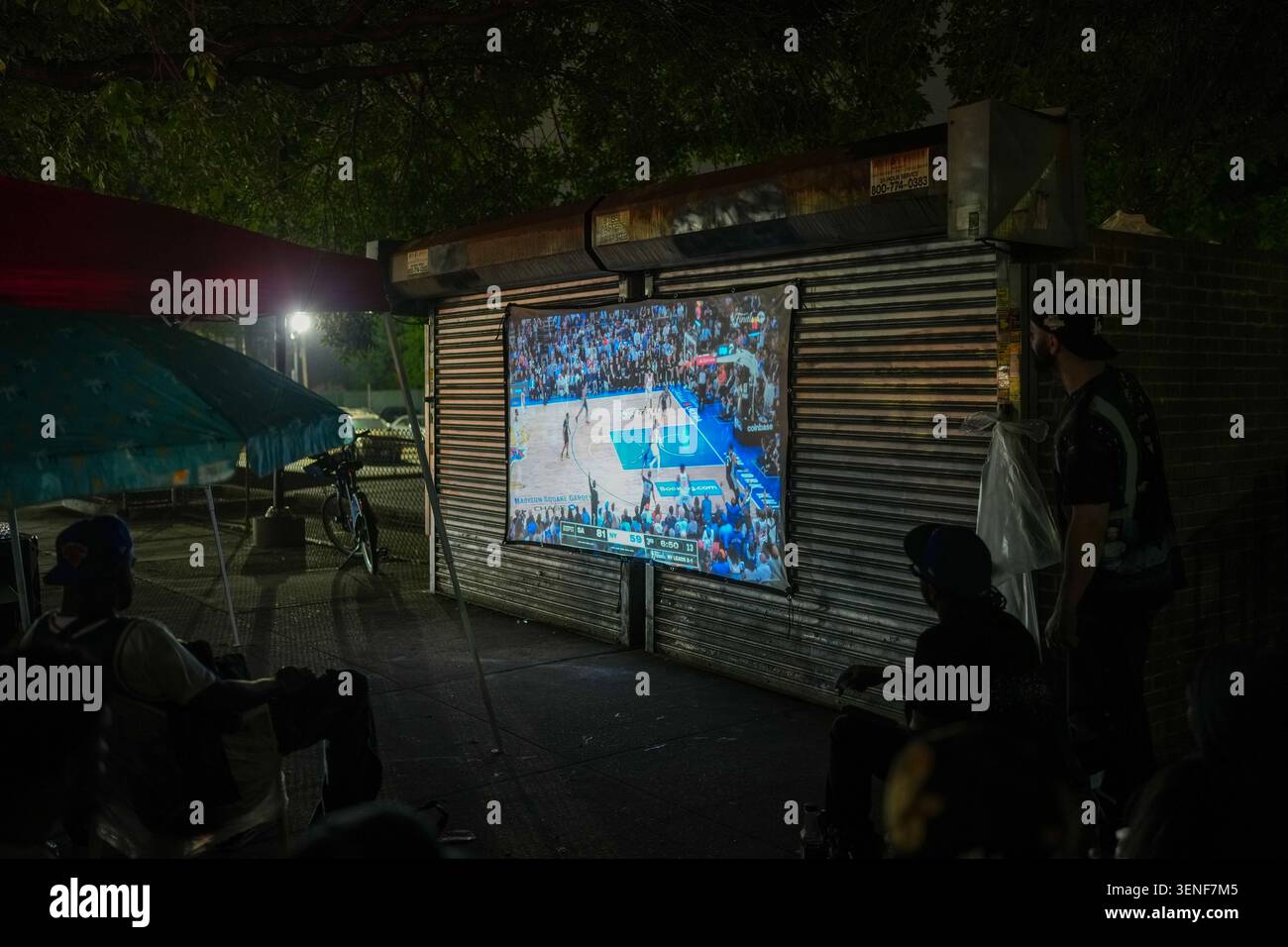 People watch a basketball game projected onto a screen in an outdoor ...