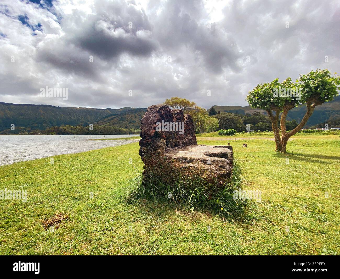 A rugged weathered stone bench sits on a green grassy lakeshore with ...
