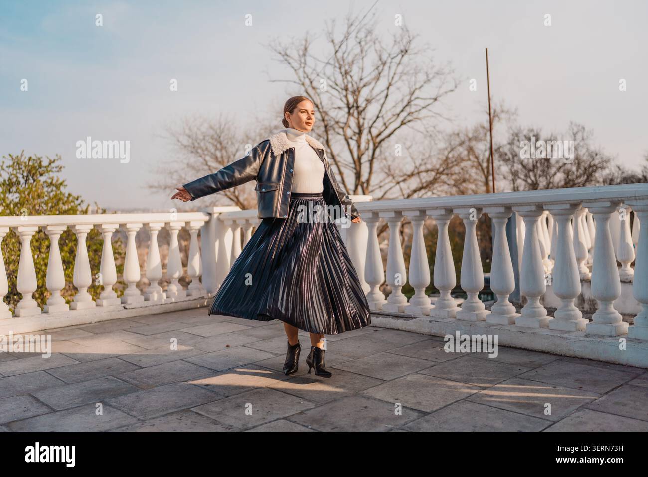 A stylish woman in a leather jacket and pleated skirt spins on a white ...