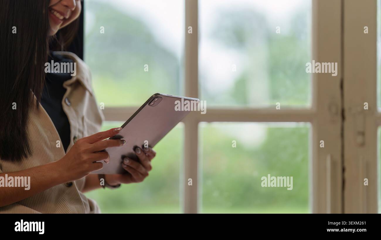 Cropped shot of smiling Asian woman using digital tablet by window ...