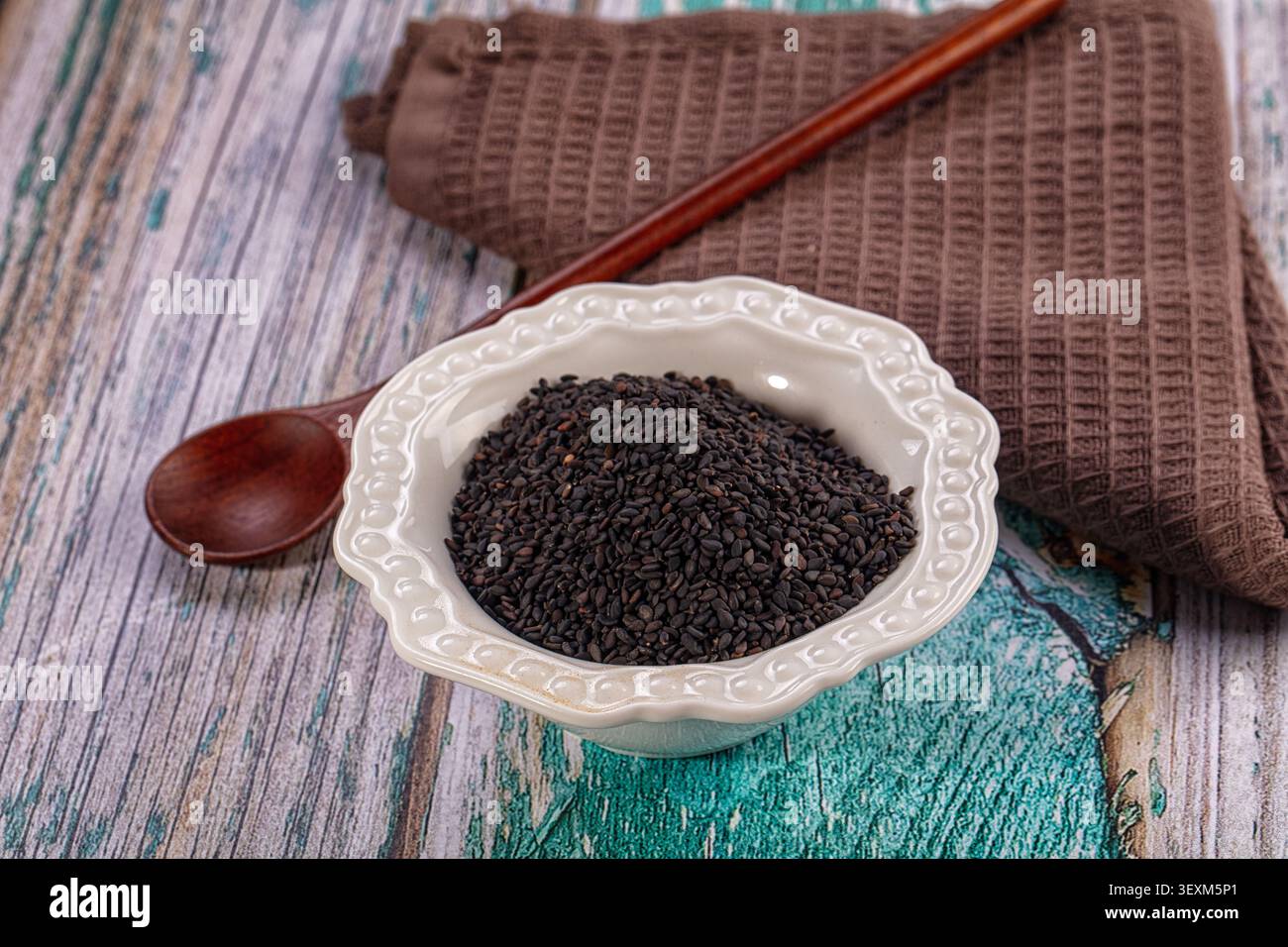 Black sesame seeds fill ornate white ceramic bowl beside wooden spoon ...