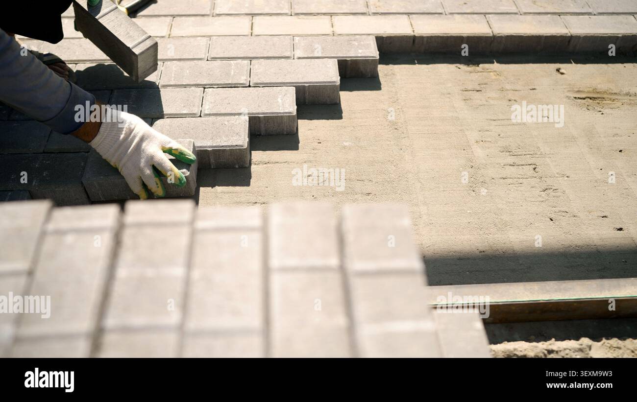Hand placing interlocking pavers on prepared sand bed. Sunlight casts ...