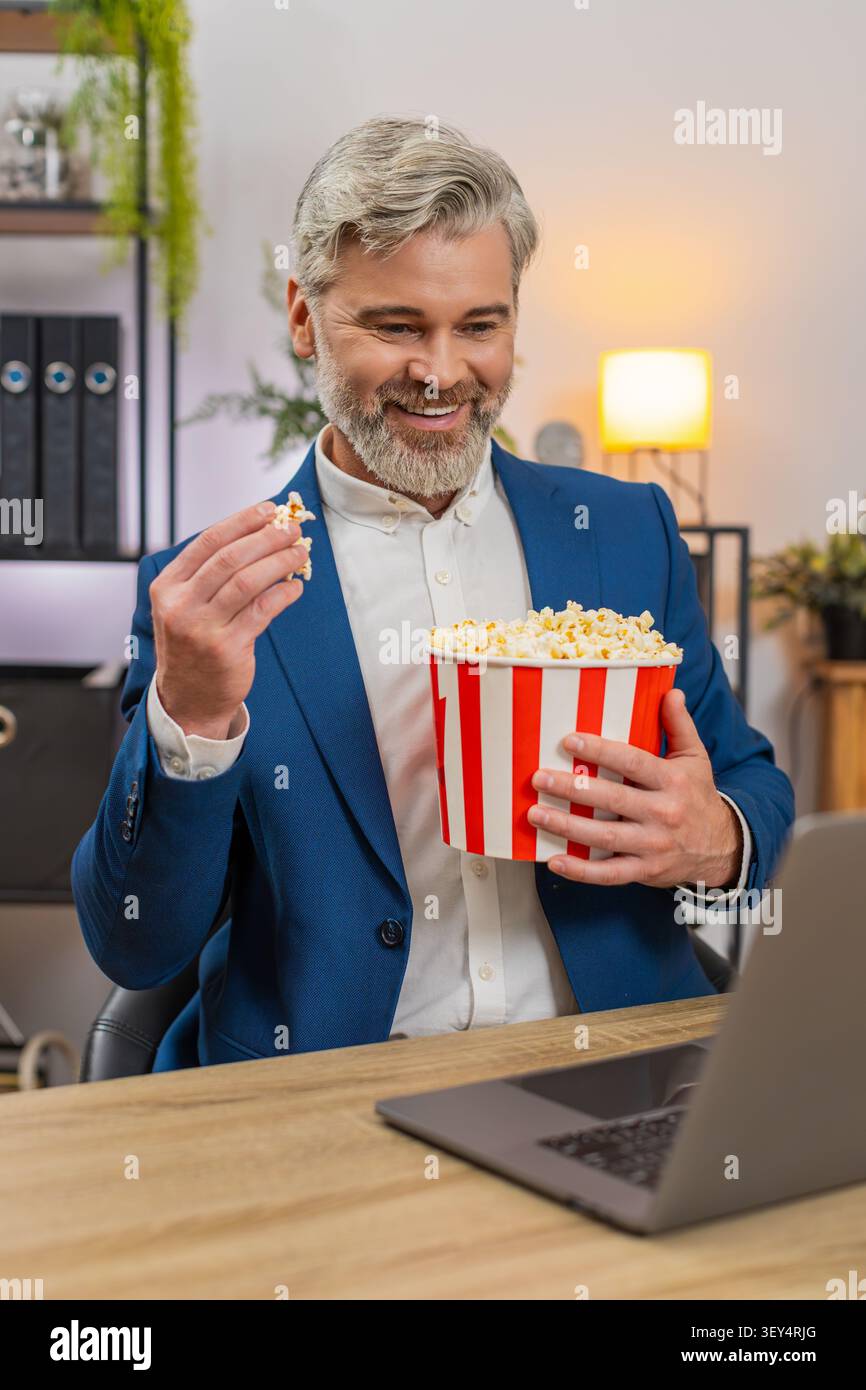 Middle-aged businessman at home office munches popcorn watching laptop ...