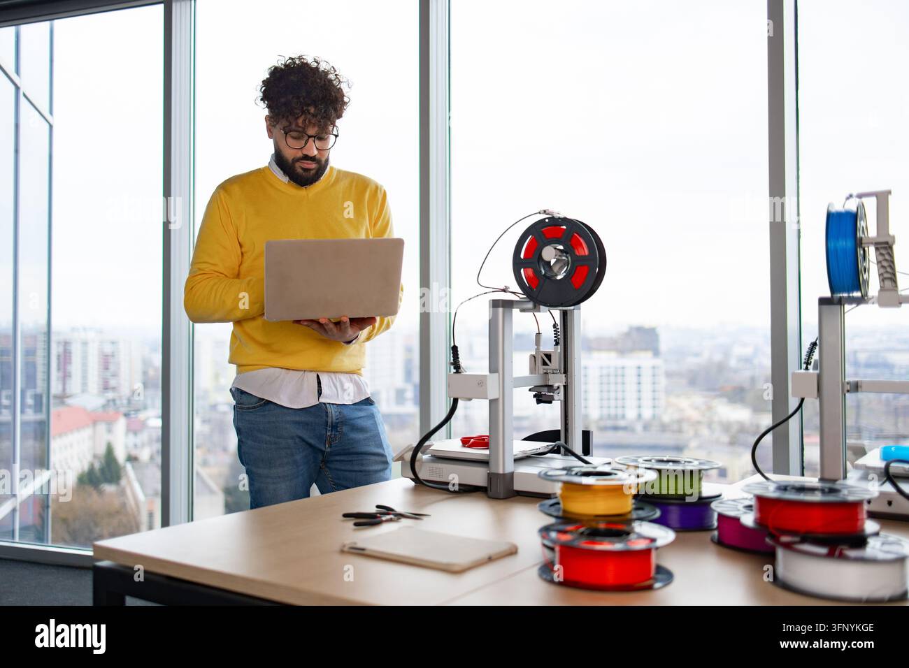 A man in a yellow sweater stands by a large window holding a laptop ...