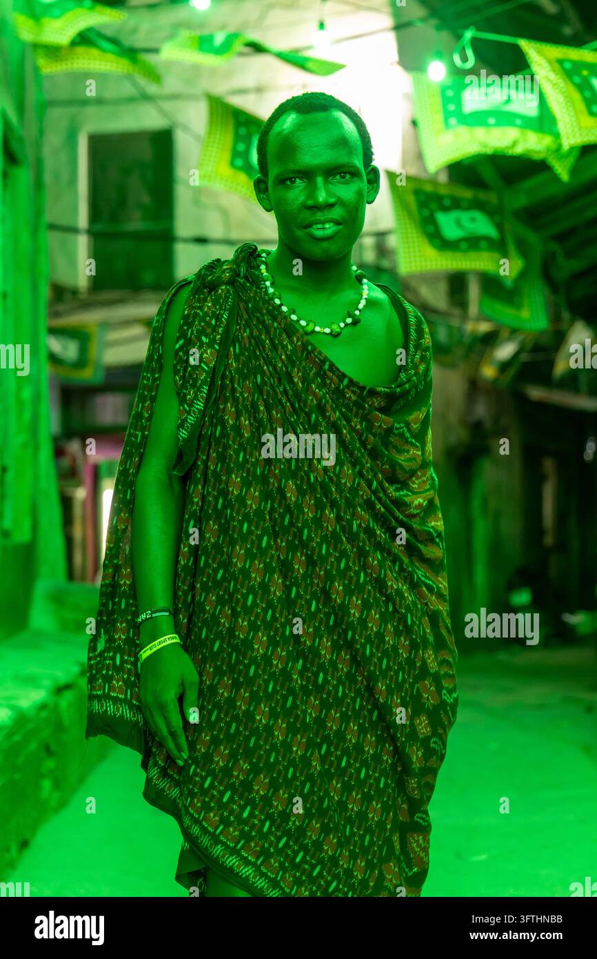 Young man in a patterned wrap and bead necklace smiling in an alley lit ...