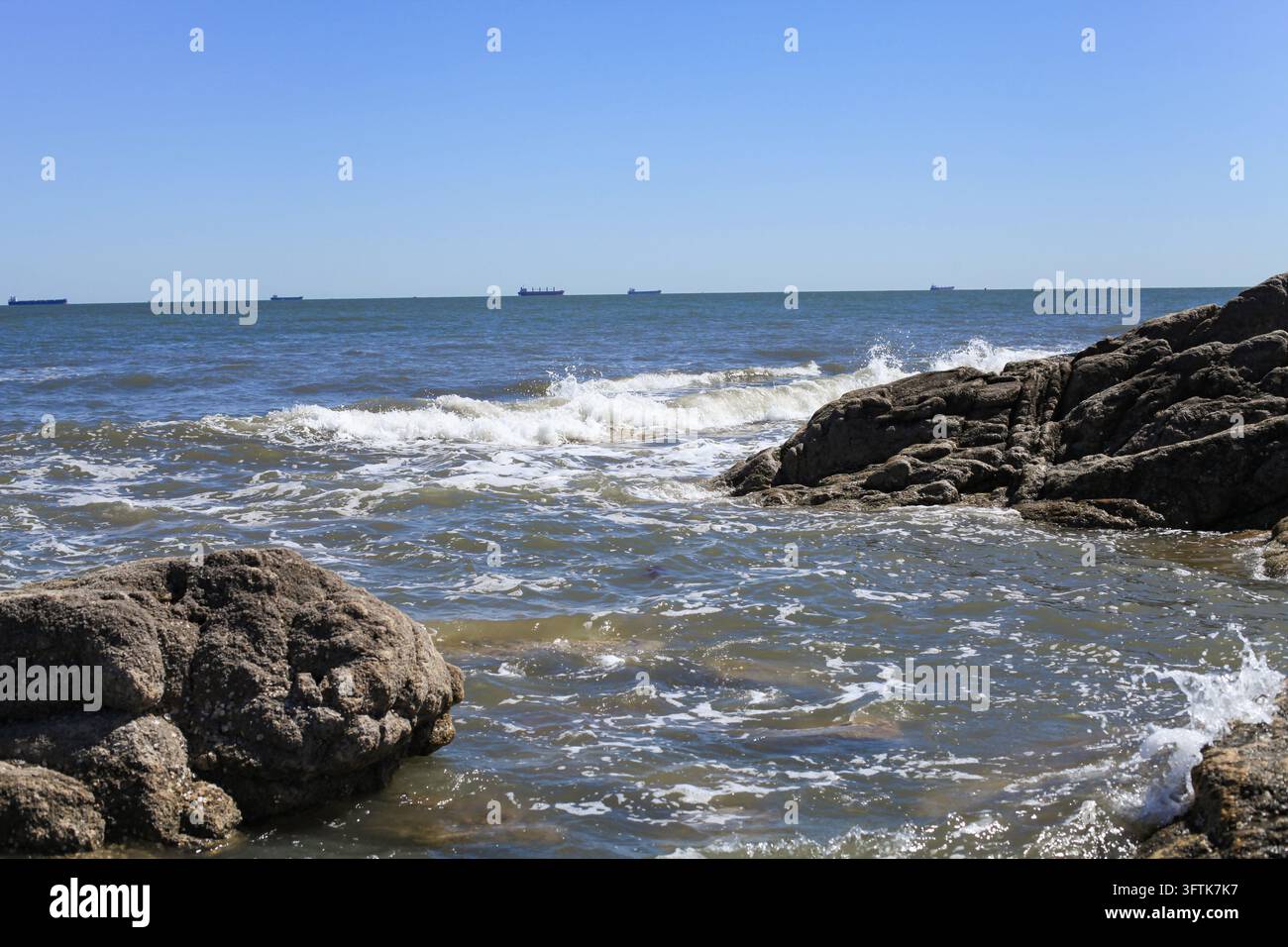 Under a clear blue sky, waves surge and crash against coastal rocks ...
