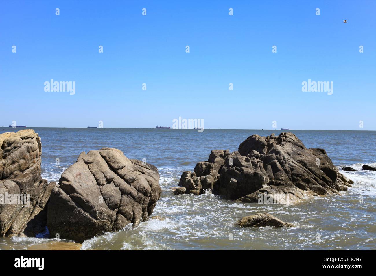 Under a clear blue sky, gentle waves lap against coastal rocks, with ...