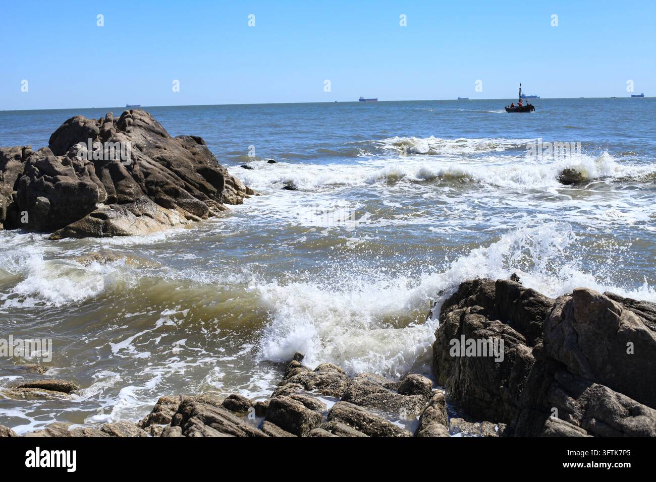 Under a clear blue sky, surging waves crash against coastal rocks ...
