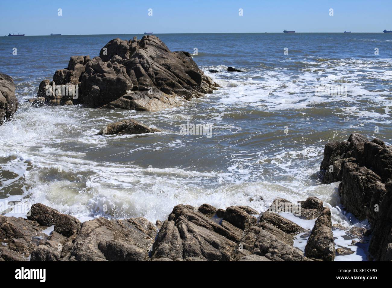 Under a clear blue sky, waves continuously crash against coastal rocks ...