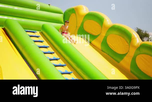 A young boy in red swim shorts slides down a vibrant green and yellow ...