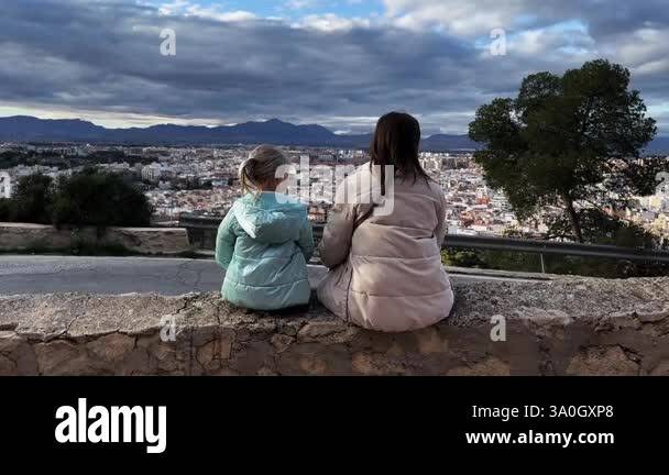 Rear view of mother and daughter tourists sitting on the parapet ...