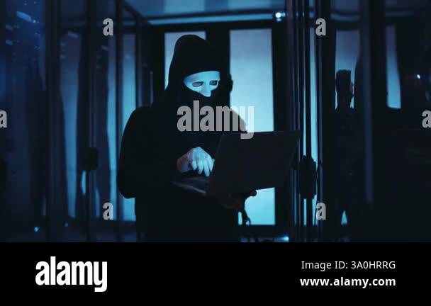 A hooded figure in a mask hacks a sensitive server room with a laptop ...