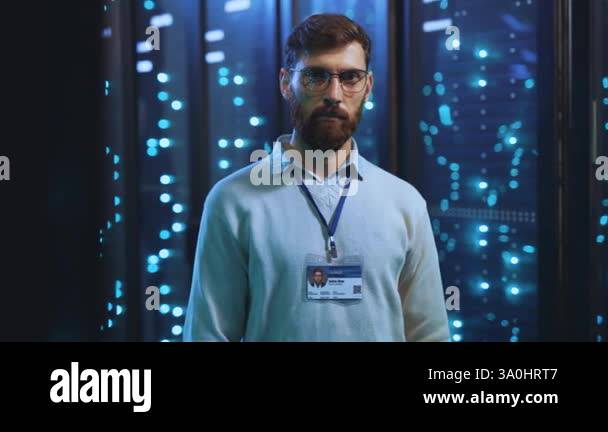 A confident bearded man with glasses stands in a hightech server room ...