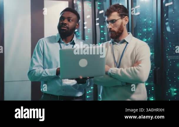 In a server room lit by blinking servers, two men discuss a critical project while examining a ...