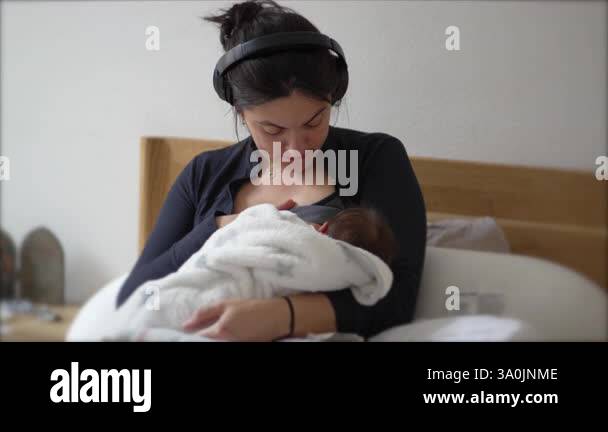 Mother wearing headphones while breastfeeding her newborn baby, sitting ...
