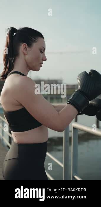 A fit female boxer takes a break, leans on a bridge fence with boxing ...