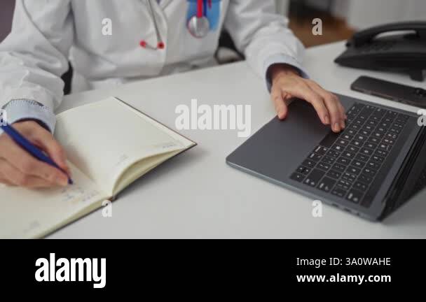 Doctor writing notes in notebook while using laptop in hospital office ...