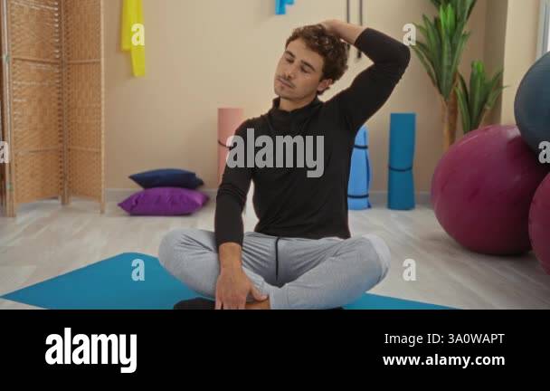 Man stretching neck in gym sitting on mat, surrounded by yoga equipment ...