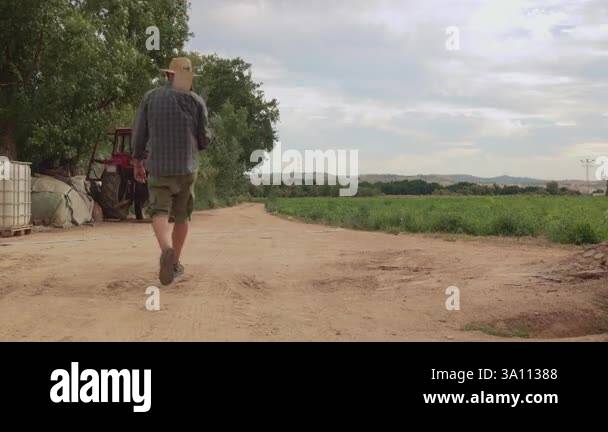 A farmer wearing a straw hat and casual work clothes walks along a dirt ...