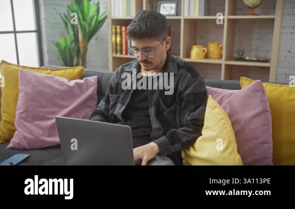 A focused hispanic man with glasses uses a laptop and smartphone in a ...