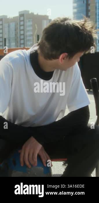 Vertical shot of young gen Z BMX rider sitting in skate park, watching ...