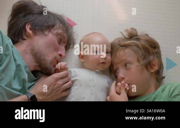 A father and elder brother gently kiss their newborn baby as they all ...
