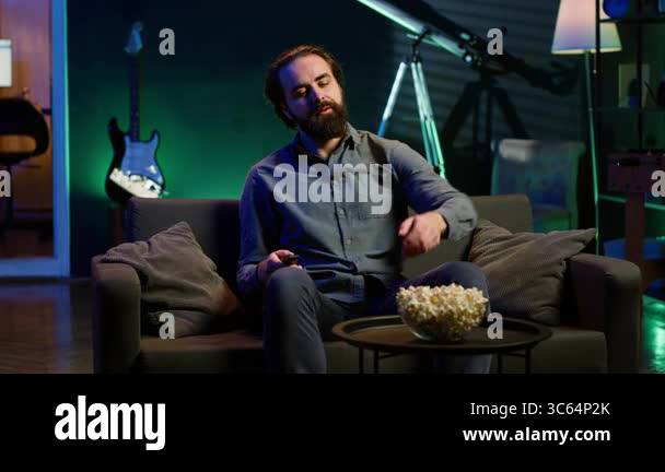 Man turning on TV using remote control, enjoying leisure time at home, munching on popcorn ...