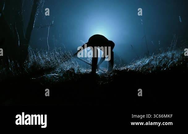 A hooded figure digs into the ground at night, illuminated by an eerie ...