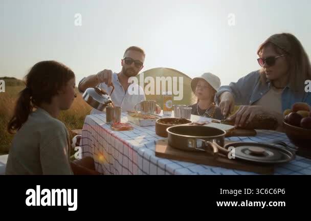 Family sitting at picnic table during camping trip, father pouring tea ...