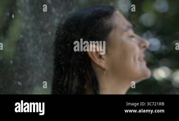 Woman with closed eyes under outdoor shower, water droplets in slow ...