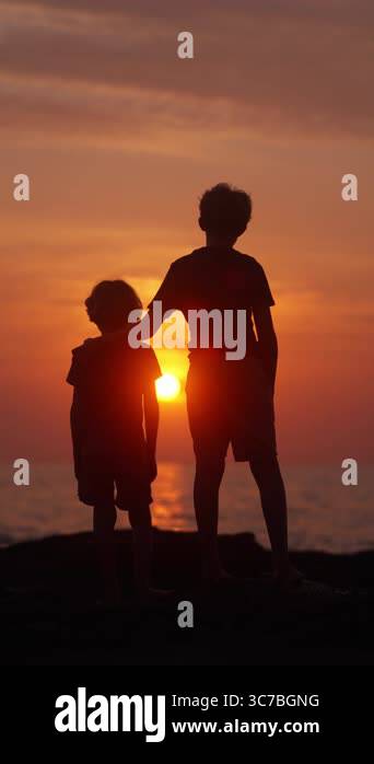 Two brothers stand side by side in silhouette as the sun sets over the ...