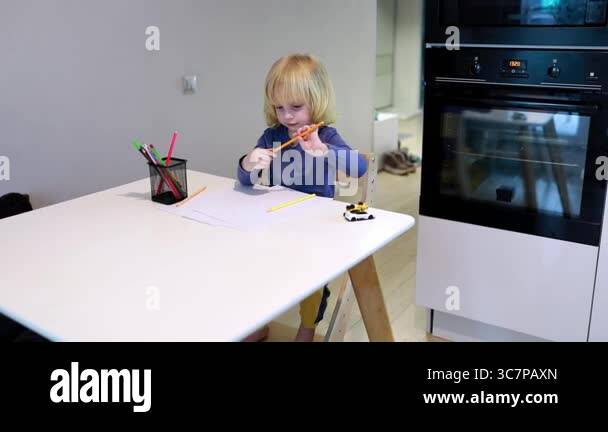 A young child sits at a white table in a cozy kitchen, drawing with ...