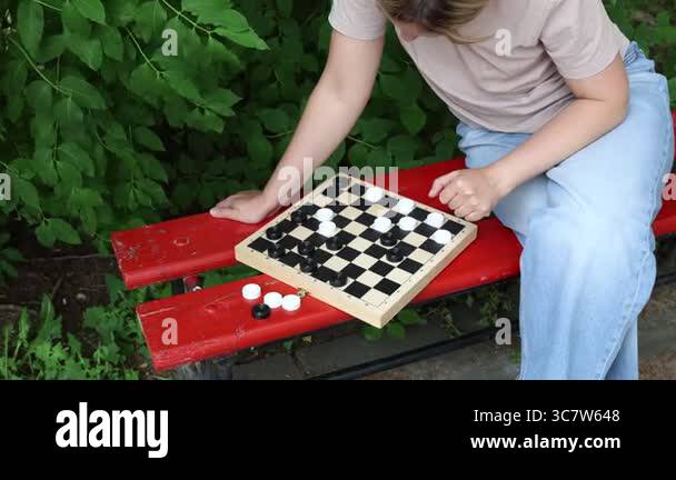 A woman sits on a red bench in a park, playing checkers with an unseen ...