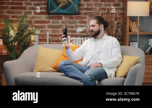 Peaceful Caucasian man sitting comfortably on sofa holding smartphone ...
