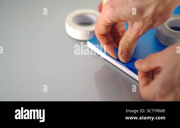 A hand secures a book cover with blue tape on a table, surrounded by ...