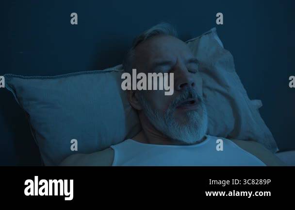 Sleepless senior man with grey hair and beard lying in bed in dimly lit ...