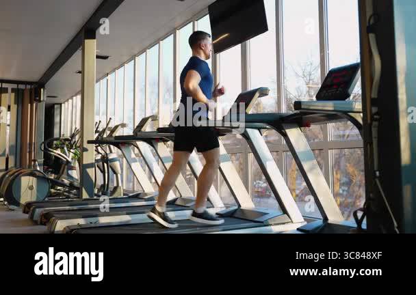 Young man jogging on a treadmill inside a bright, modern gym with large ...