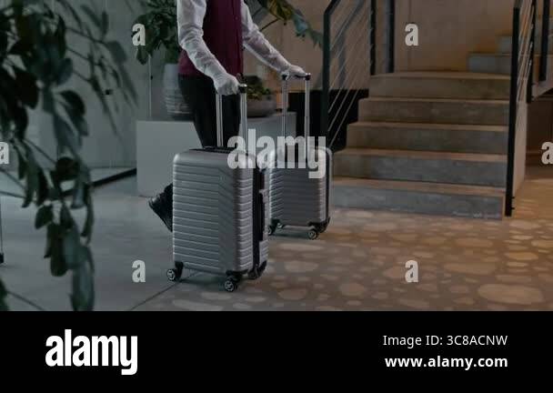 Hotel bellhop wearing gloves and uniform moving two suitcases across ...