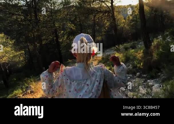 Young blonde female adjusting backpack straps while walking along forest path during golden ...
