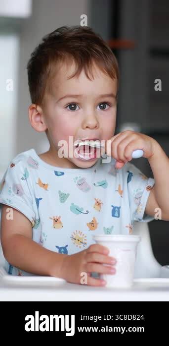 Lovely toddler sits at high table eating yoghurt from plastic cup. Baby ...