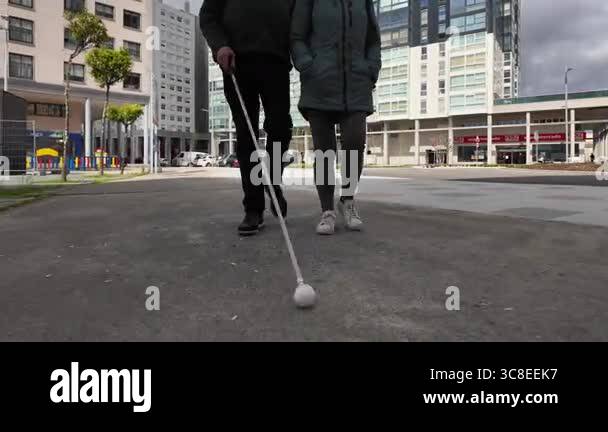 Blind man walking on the pavement using his cane and a woman providing ...