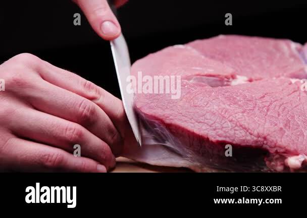 A butcher removes the fascia from a beef steak with a large knife in a ...