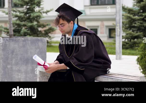 Young man sits on bench gazing thoughtfully at diploma. Student looks ...