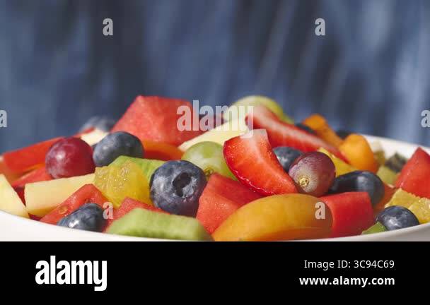 bowl of fresh fruit salad rotating on blue background, food closeup ...