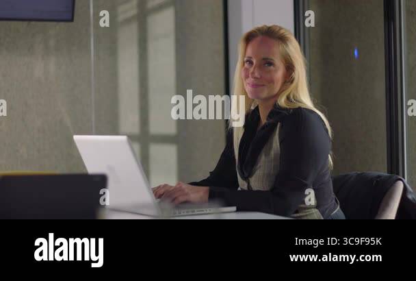 Smiling business woman seated at desk looking up from laptop with calm ...
